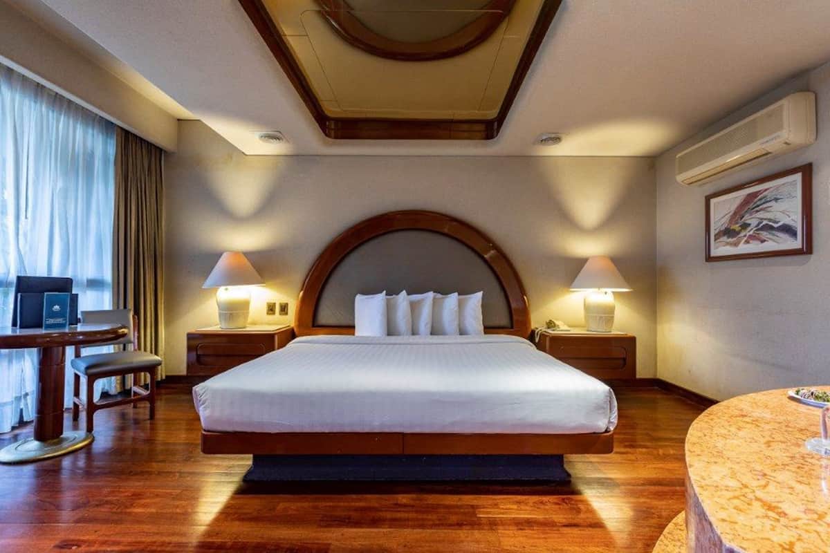 Quarto de hotel com uma cama grande, duas mesas de cabeceira com luminárias, uma escrivaninha com cadeira e uma mesa redonda com um copo.