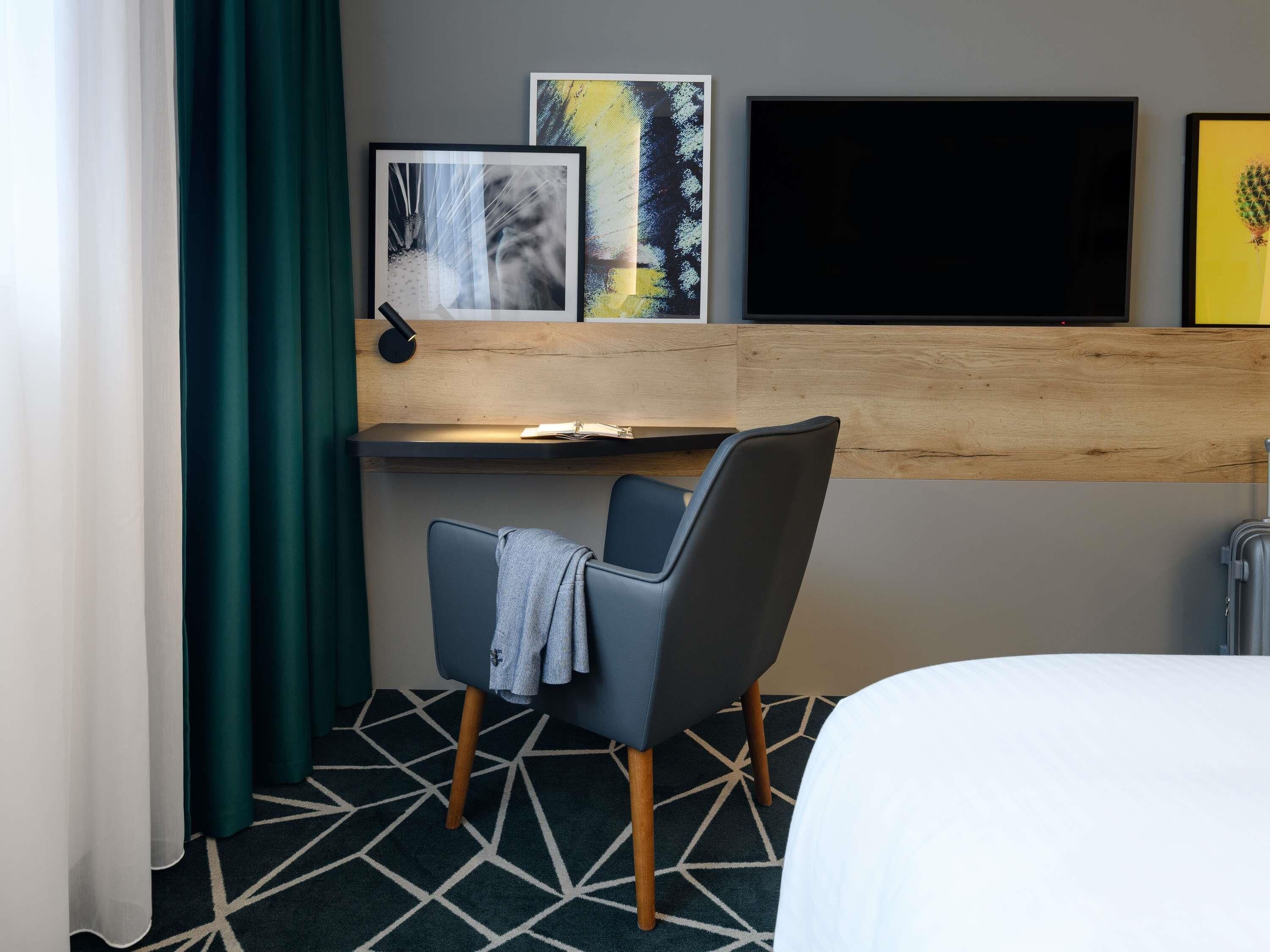 Quarto de hotel com cama, escrivaninha com cadeira, televisão e obras de arte na parede.