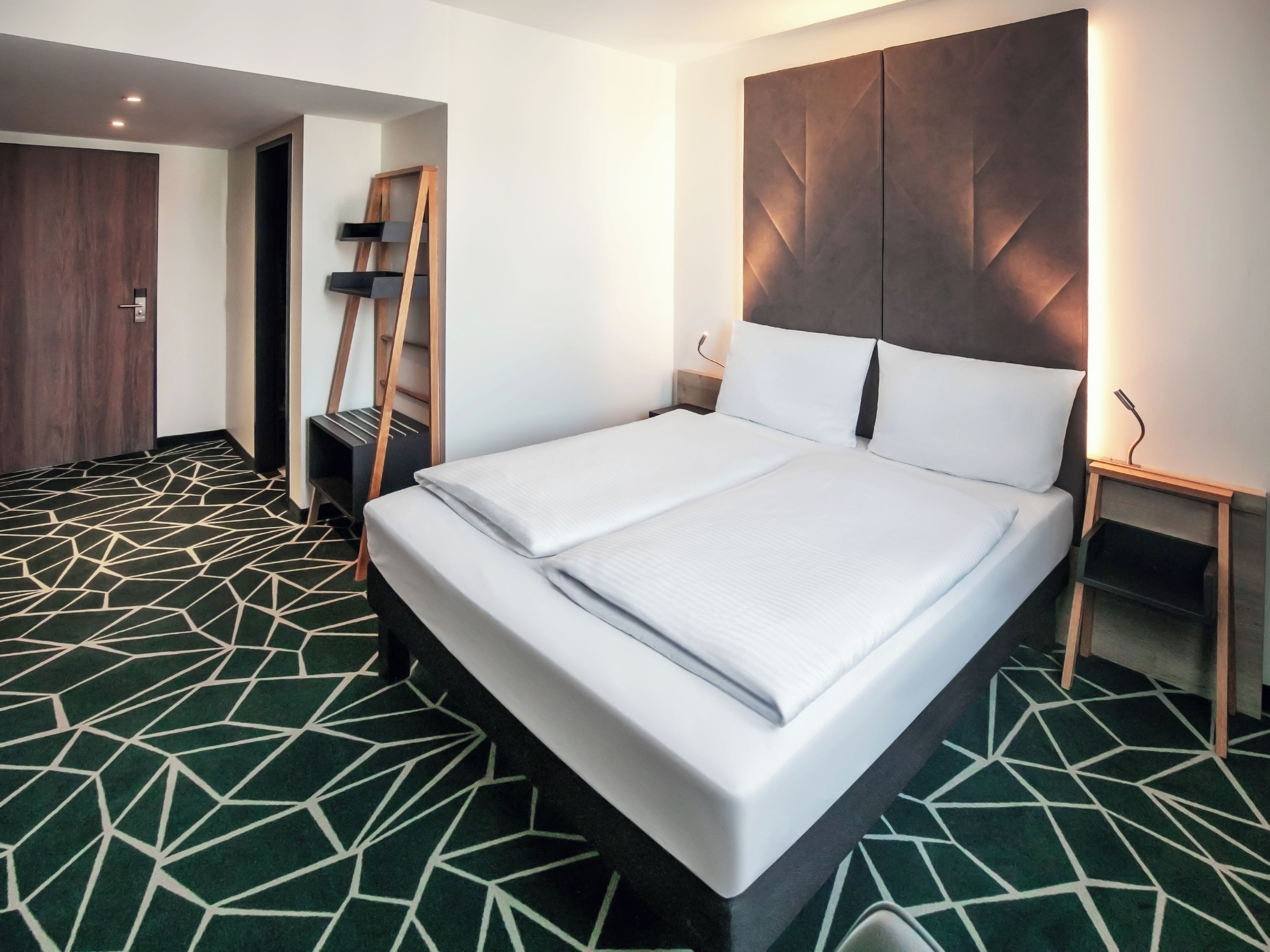 Quarto de hotel com uma cama grande, cabeceira de madeira, mesa de cabeceira e tapete com padrão geométrico.
