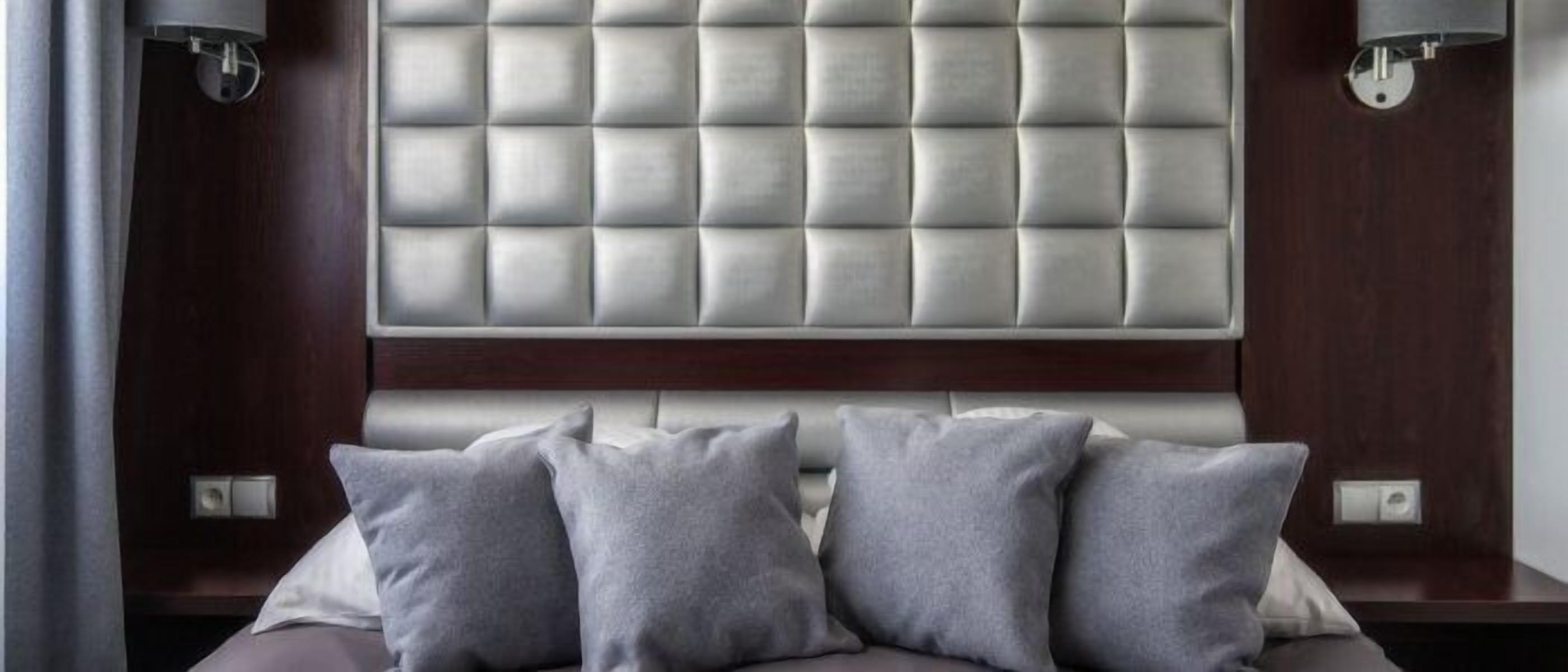 Quarto de hotel com cabeceira estofada, cama com roupa de cama cinzenta e luminárias fixadas na parede.