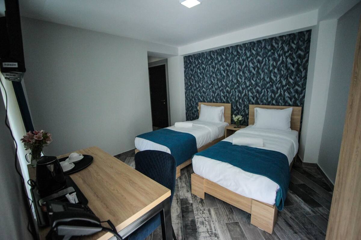 Quarto de hotel com duas camas de solteiro, escrivaninha de madeira, cafeteira e pequena mesa com um vaso de flores.