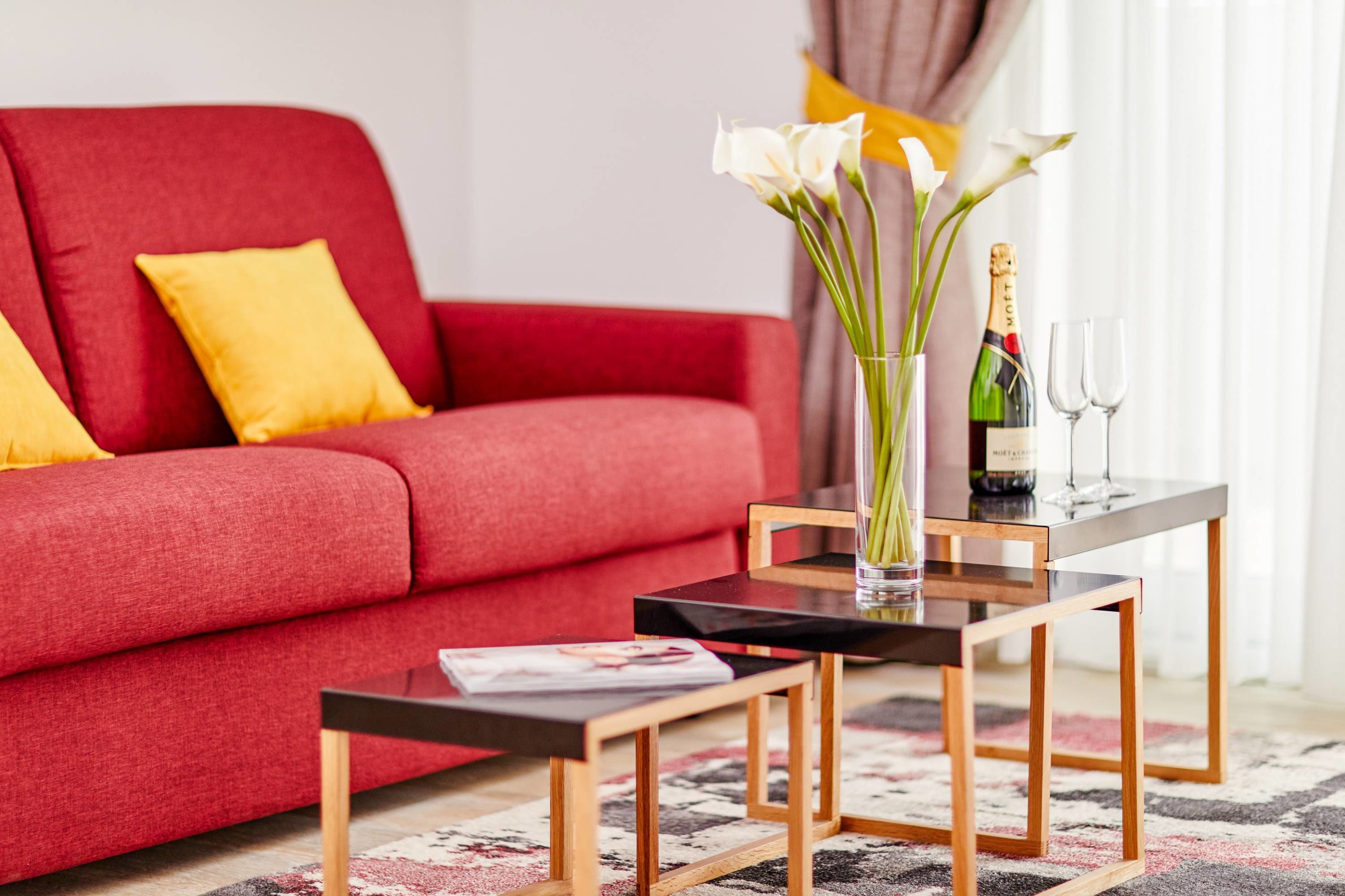 Uma sala de estar moderna com um sofá vermelho, almofadas amarelas, um vaso de vidro com flores, uma garrafa de champanhe e duas taças sobre uma mesa de centro de madeira.