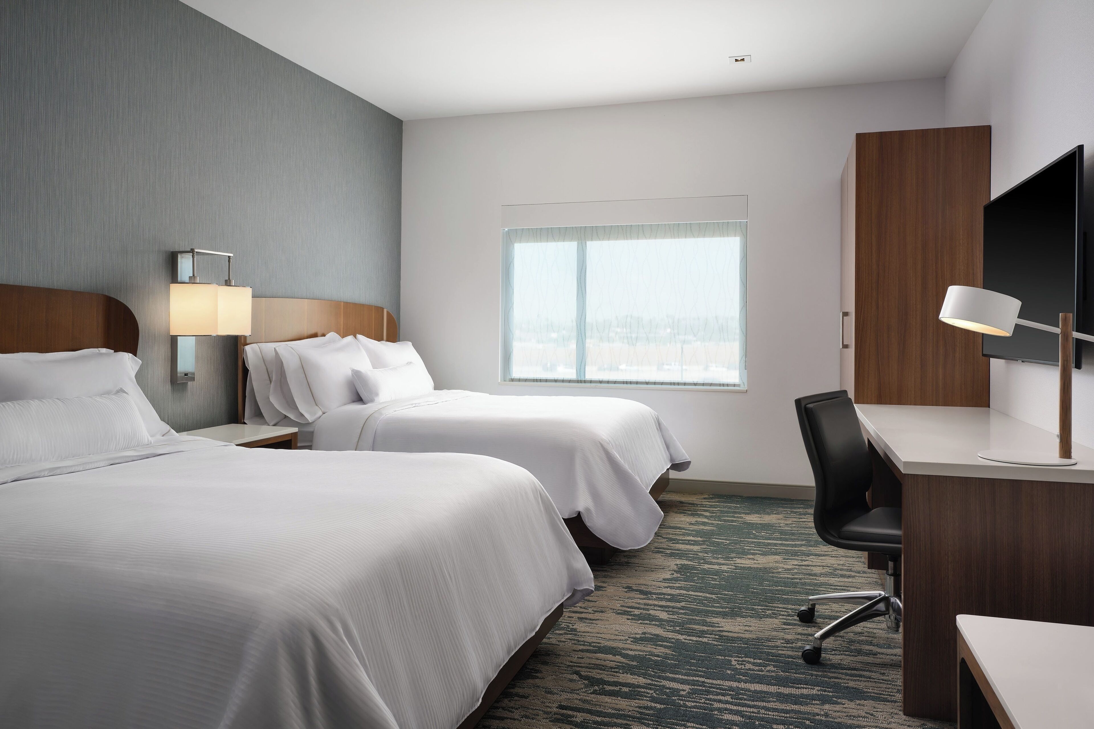 Quarto de hotel com duas camas, uma escrivaninha, uma cadeira, televisão e janela com vista.