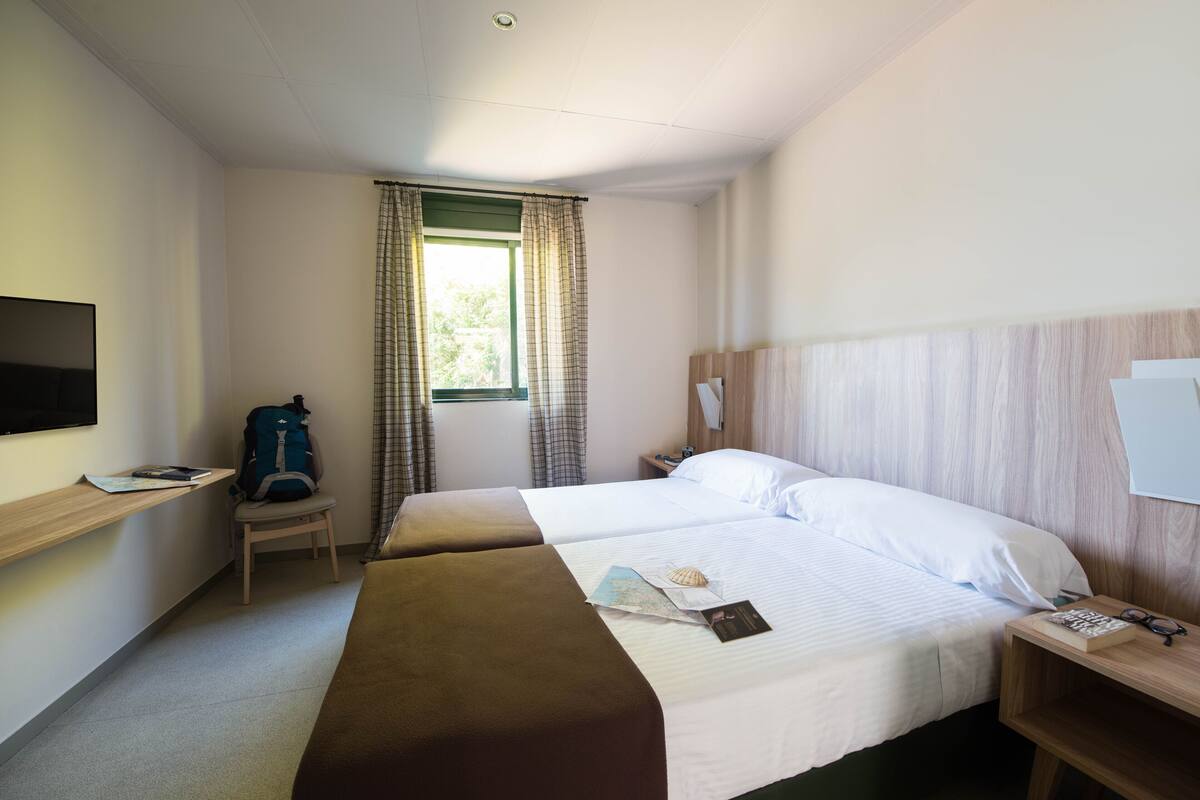 Quarto de hotel com duas camas, televisão, escrivaninha e janela com cortinas.