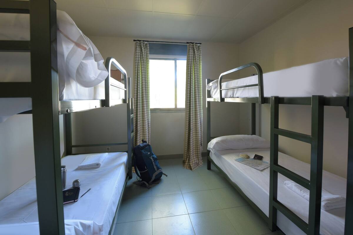 Um dormitório com beliches, uma janela com cortinas, uma mochila e uma garrafa de água no chão.