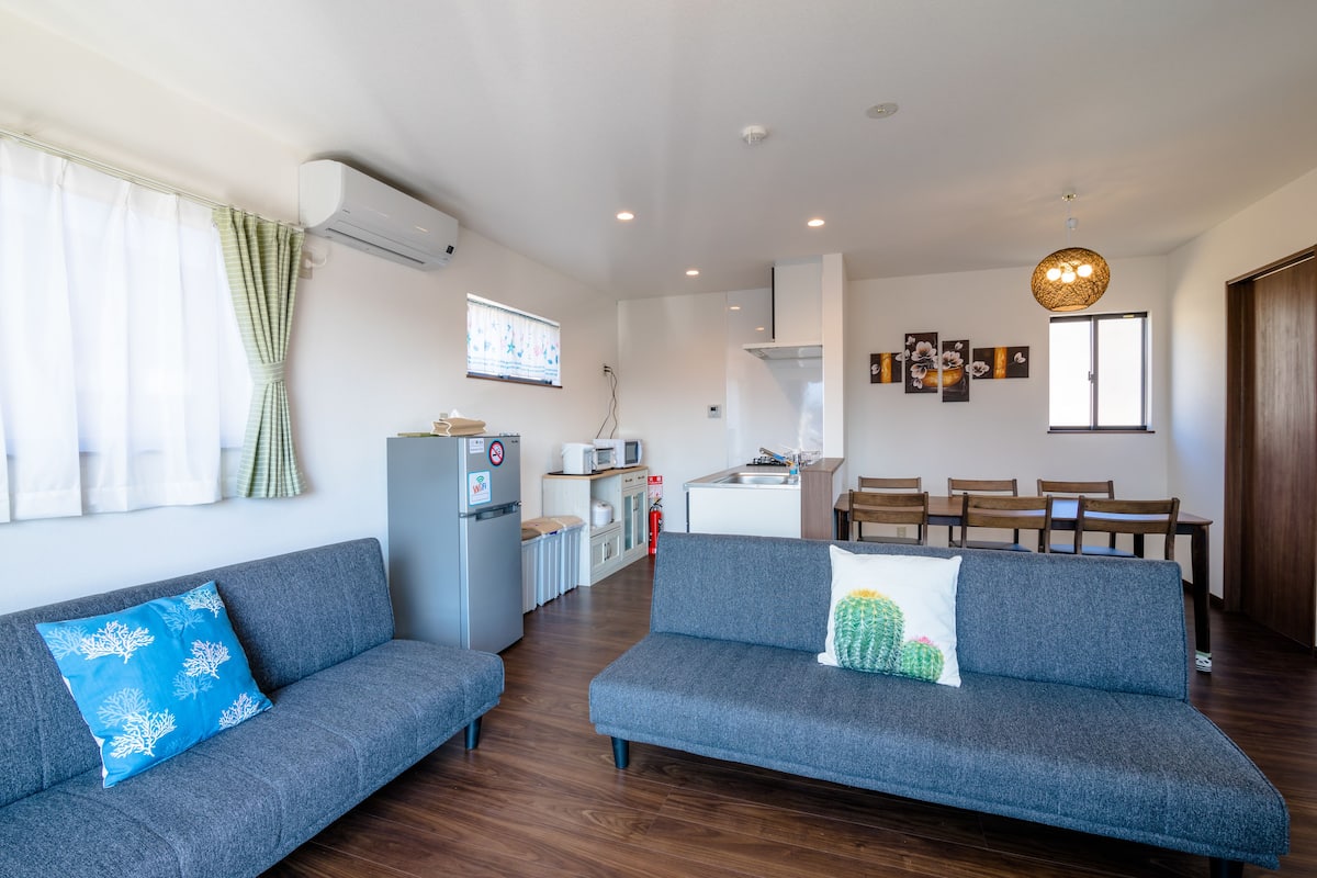 Uma sala de estar moderna com dois sofás cinzentos, uma mesa de jantar com cadeiras, uma área de cozinha e uma janela grande com cortinas.