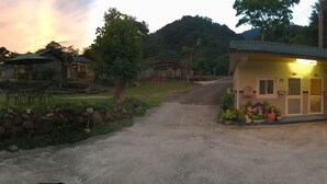 Um edifício de dois andares com um alpendre, um caminho de terra que leva à entrada e um pequeno jardim com plantas em vasos.