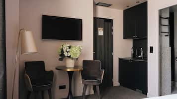 Quarto de hotel compacto com televisão de tela plana, uma pequena mesa redonda com duas cadeiras, um vaso com flores, uma área de cozinha moderna e uma porta com uma placa.