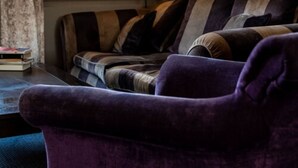 Uma poltrona roxa num quarto com um sofá riscado e uma mesa com livros.