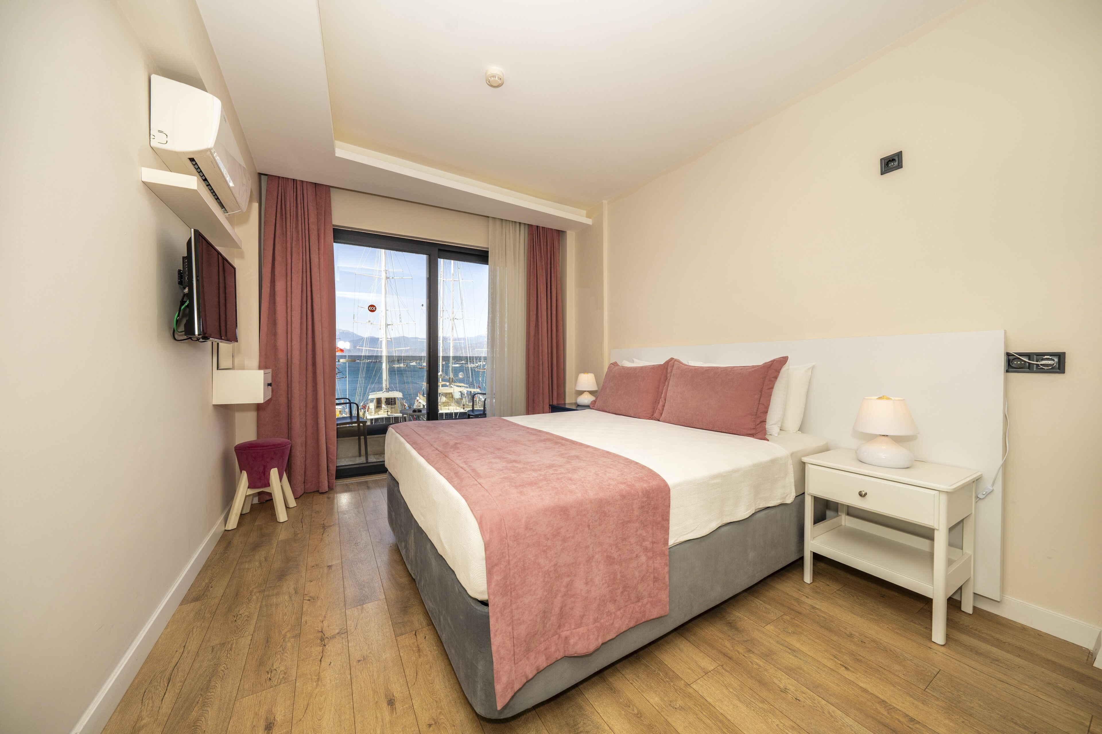 Quarto de hotel moderno com cama, mesa de cabeceira e vista para barcos pela janela.