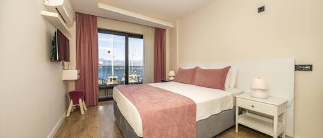 Quarto de hotel moderno com cama, mesa de cabeceira e vista para barcos pela janela.