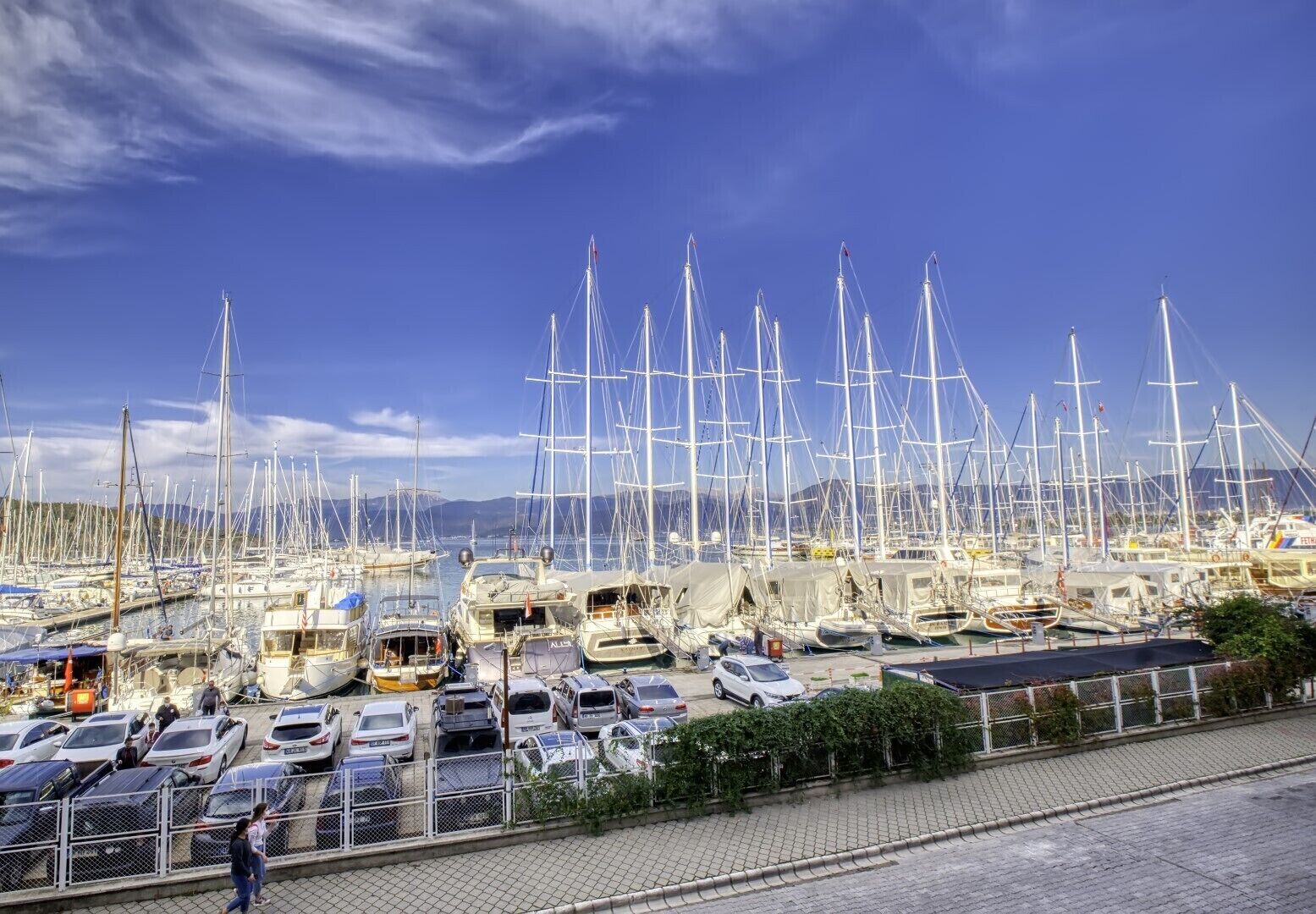 Uma marina com vários veleiros atracados, uma área de estacionamento com carros e um céu azul claro.
