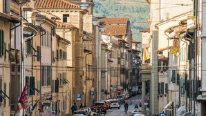 Rua com carros estacionados e edifícios, com uma estrutura em forma de cúpula ao fundo, e placas indicando direções e estacionamento.