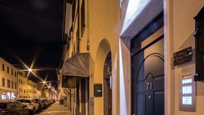 Vista noturna da rua com fileira de carros estacionados, um edifício com porta preta identificada com o número '71', e placas iluminadas que indicam uma suíte de luxo.