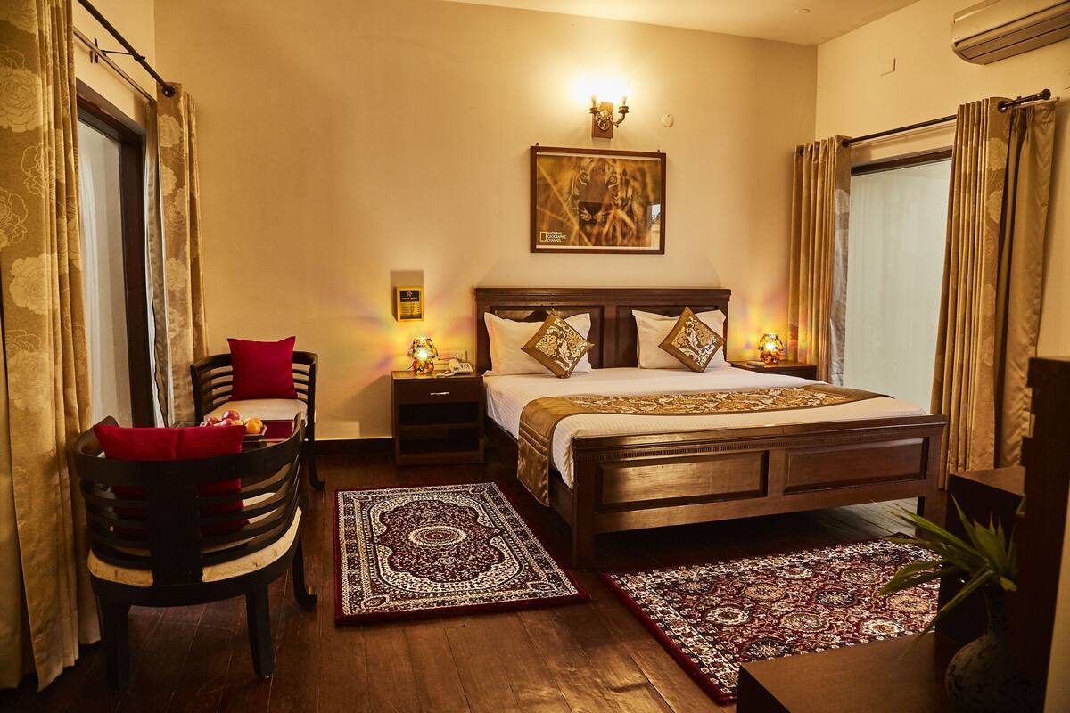 Quarto de hotel com cama, poltrona, mesa de cabeceira, luminária na parede e janela com cortinas.