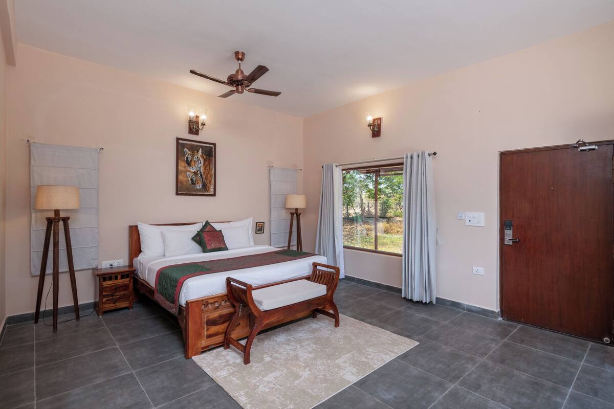 Quarto com cama, mesas de cabeceira, um banco, ventilador de teto, janela com cortinas e porta.