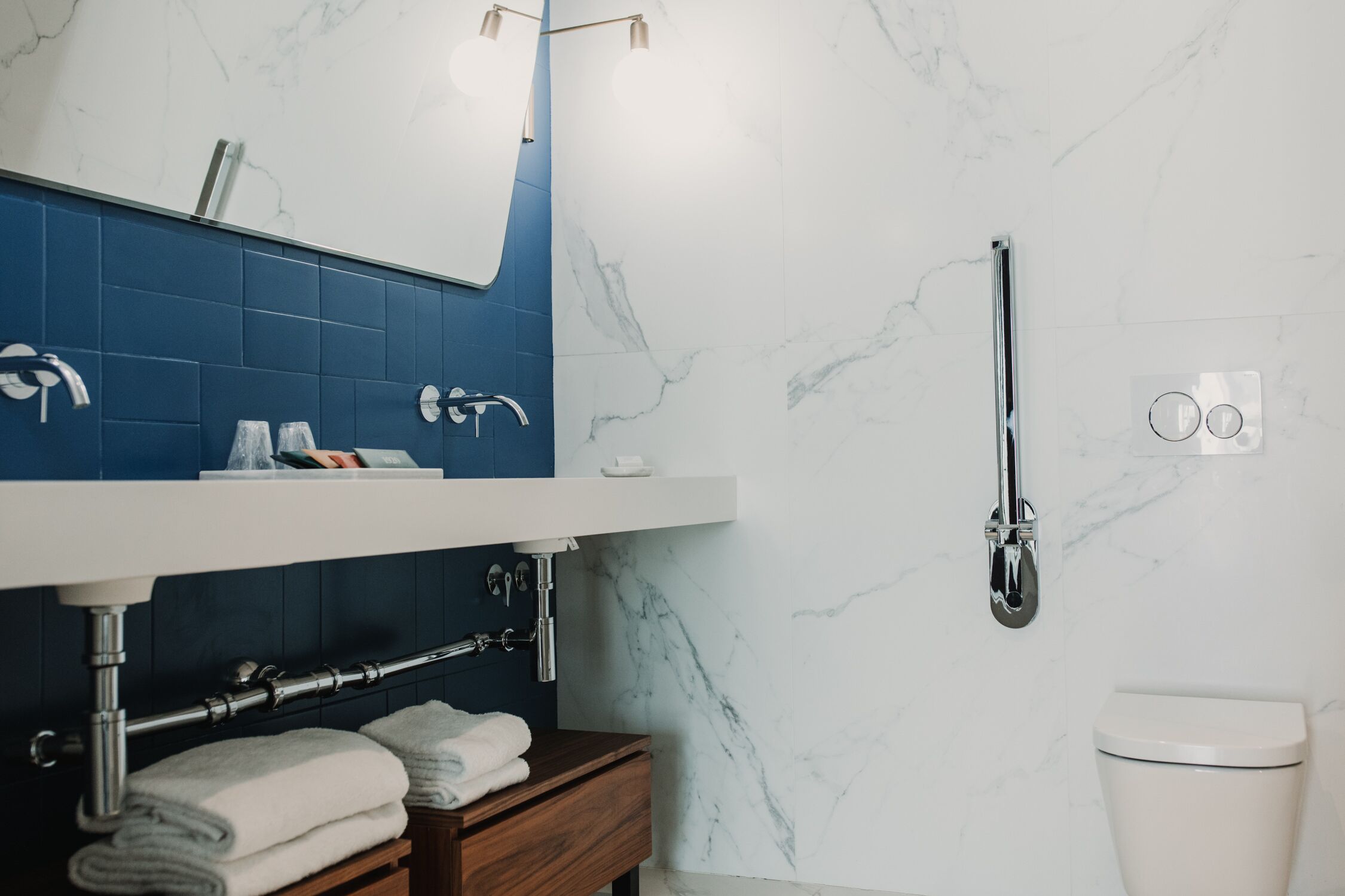Casa de banho com uma pia branca, azulejos azuis, um espelho, um porta-toalhas e uma sanita.