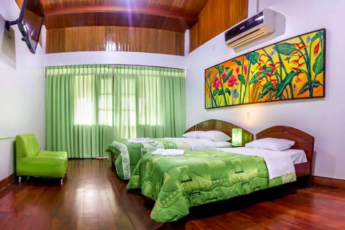 Quarto de hotel com teto curvo, cama com roupa de cama verde, poltrona verde, janela grande com cortinas verdes e um quadro colorido na parede.