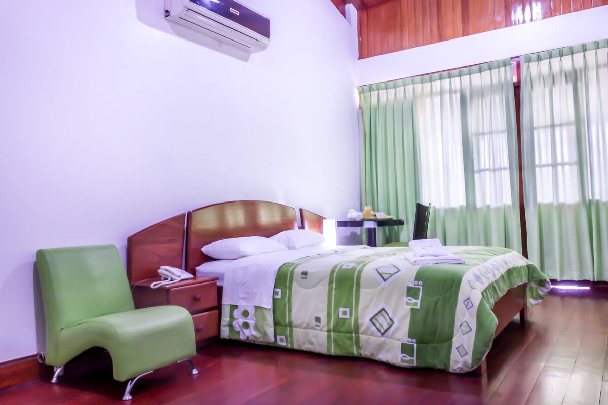Quarto de hotel com cama de madeira, poltrona verde, mesa de cabeceira com telefone e janela com cortinas verdes.