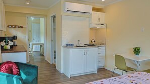 Cozinha compacta com armários brancos, bancada e pia. Há uma área de jantar com mesa e cadeiras, e uma cama com colcha estampada.