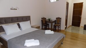 Uma cama bem arrumada com roupa de cama branca, uma pequena mesa com cadeiras e uma porta ao fundo.