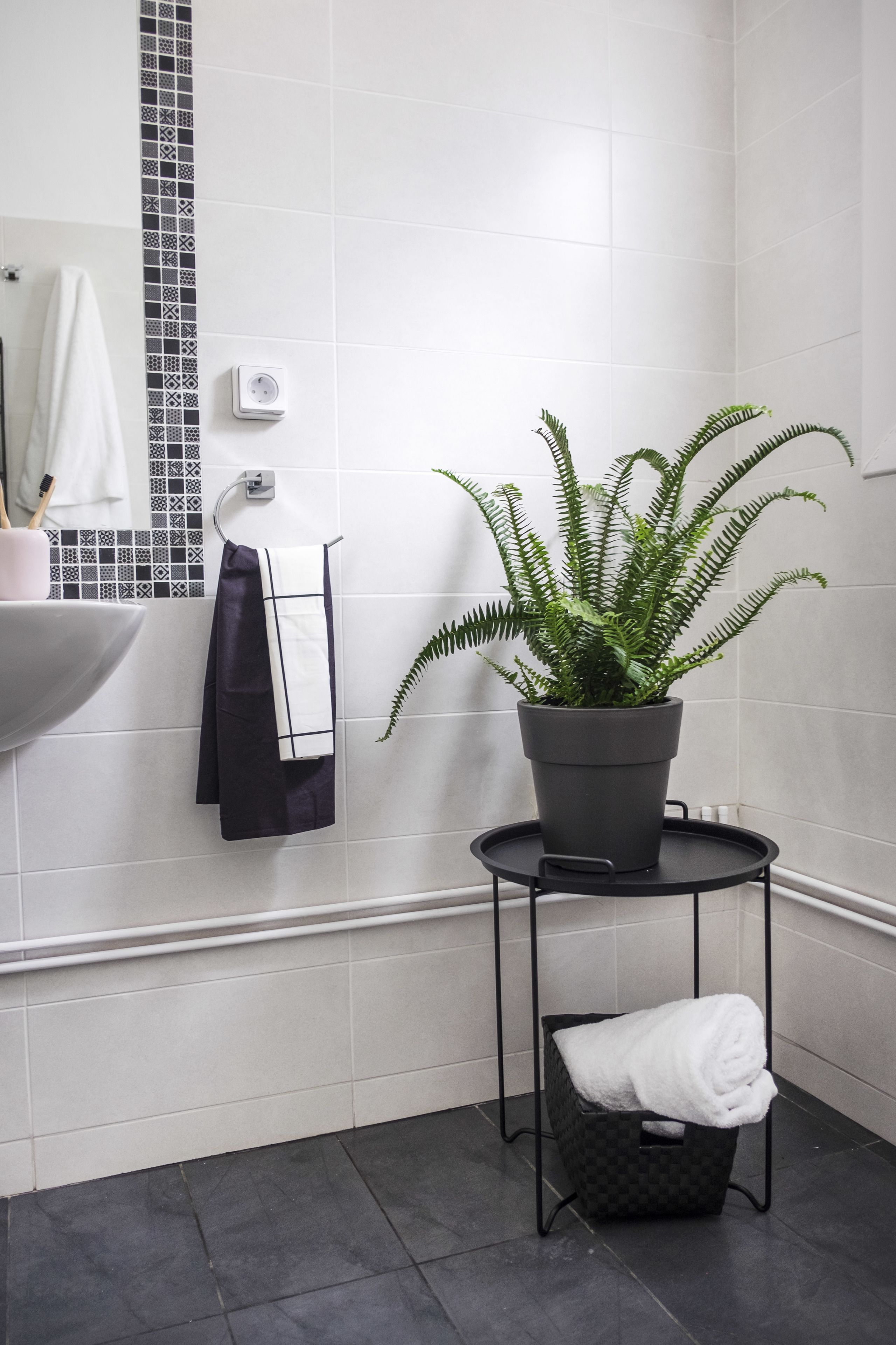 Casa de banho com azulejos brancos, faixa de azulejos com padrão preto e branco, lavatório branco, toalheiro preto, samambaia em vaso e uma mesa redonda preta com toalha branca.
