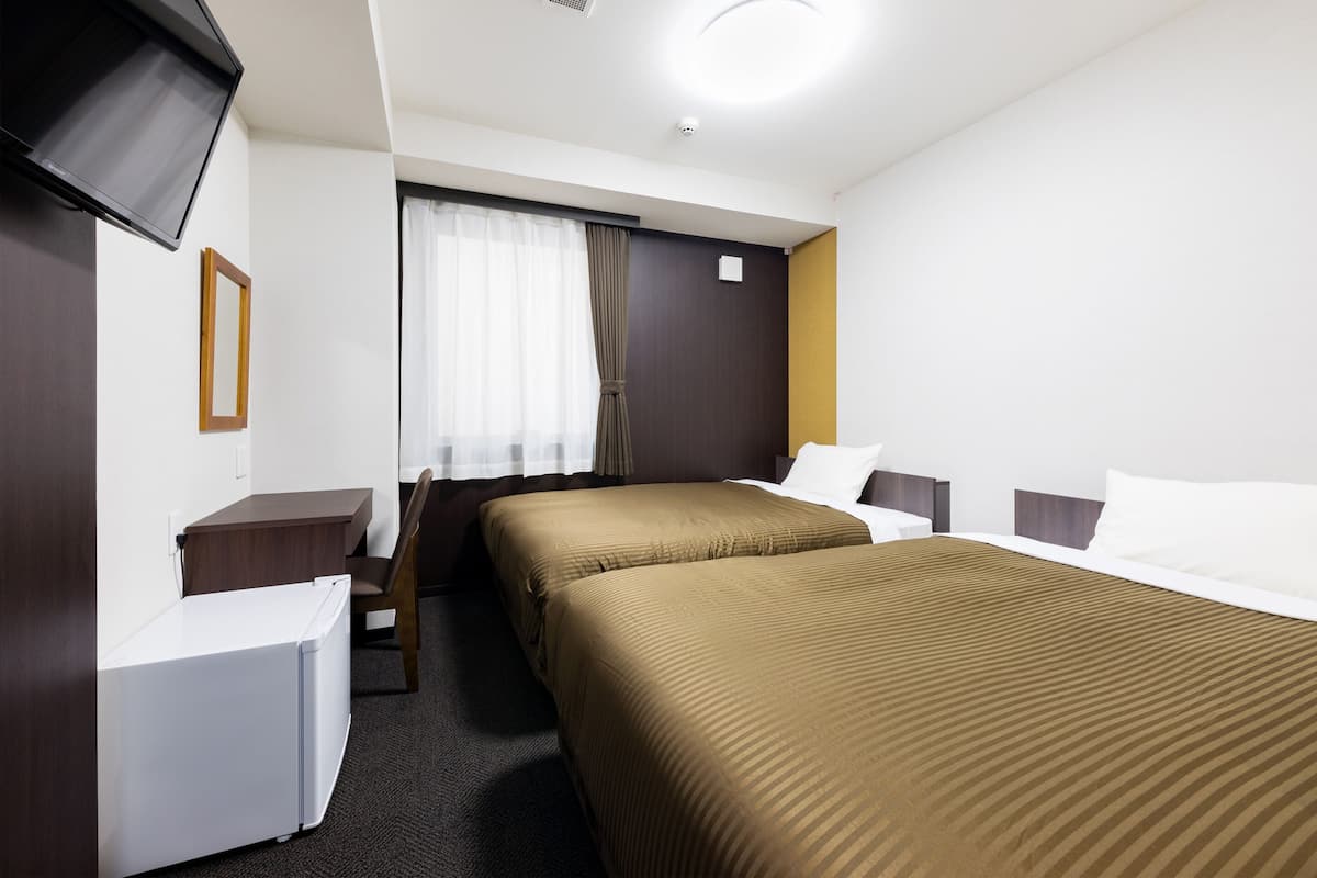 Quarto de hotel com duas camas, uma TV na parede, uma pequena mesa de trabalho e uma janela com cortinas.