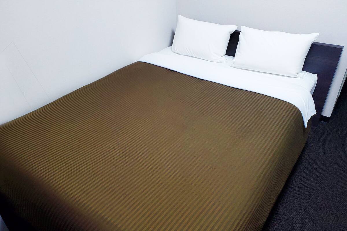 Uma cama bem arrumada com colcha marrom e almofadas brancas, cabeceira escura e parede branca lisa.