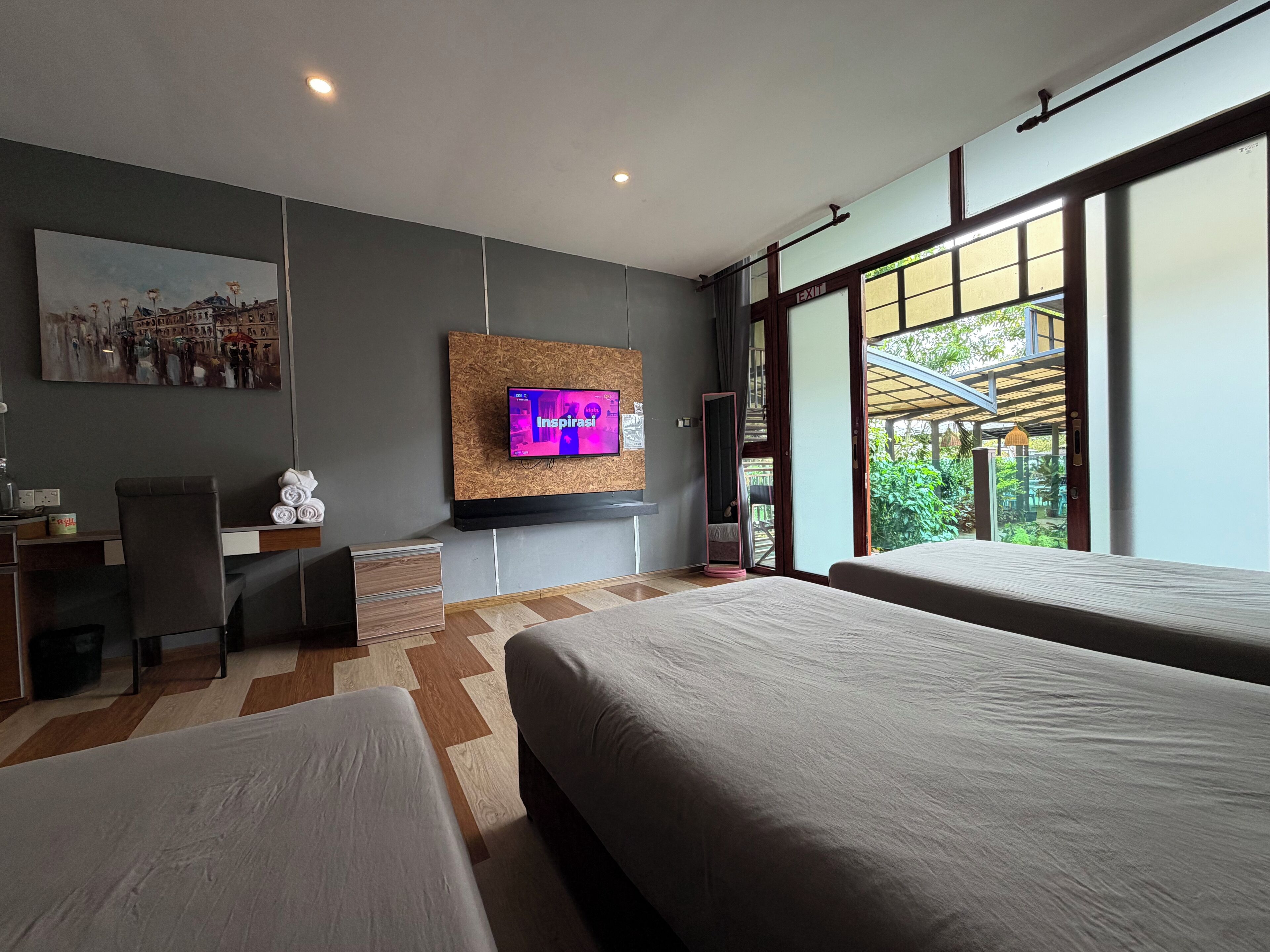 Quarto de hotel moderno com duas camas, uma escrivaninha com computador, televisão de tela plana fixada em um painel de madeira e janelas amplas com vista para o verde lá fora.