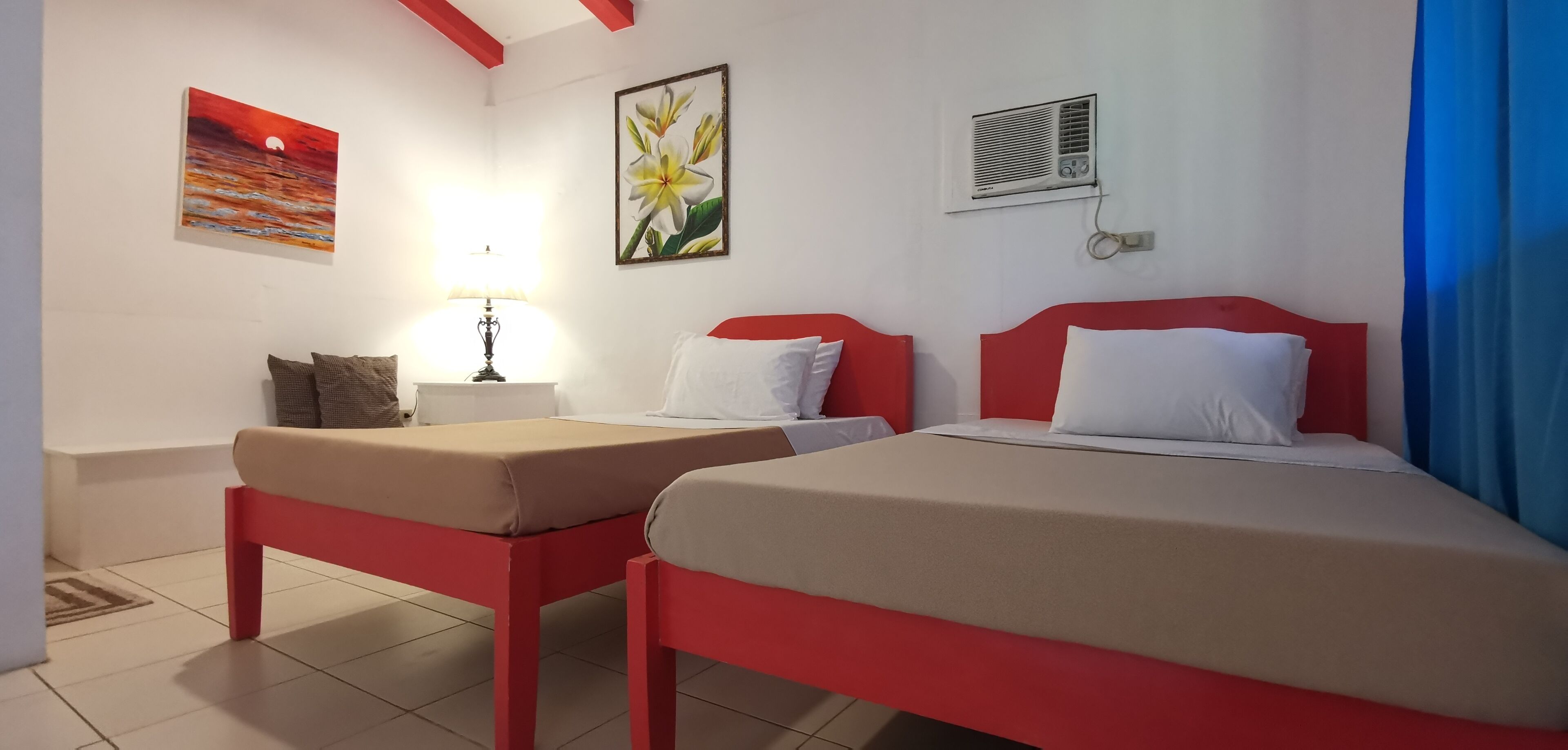 Quarto de hotel com duas camas, uma mesa de cabeceira, abajur, pintura de um pôr do sol, estampa floral e ar condicionado.