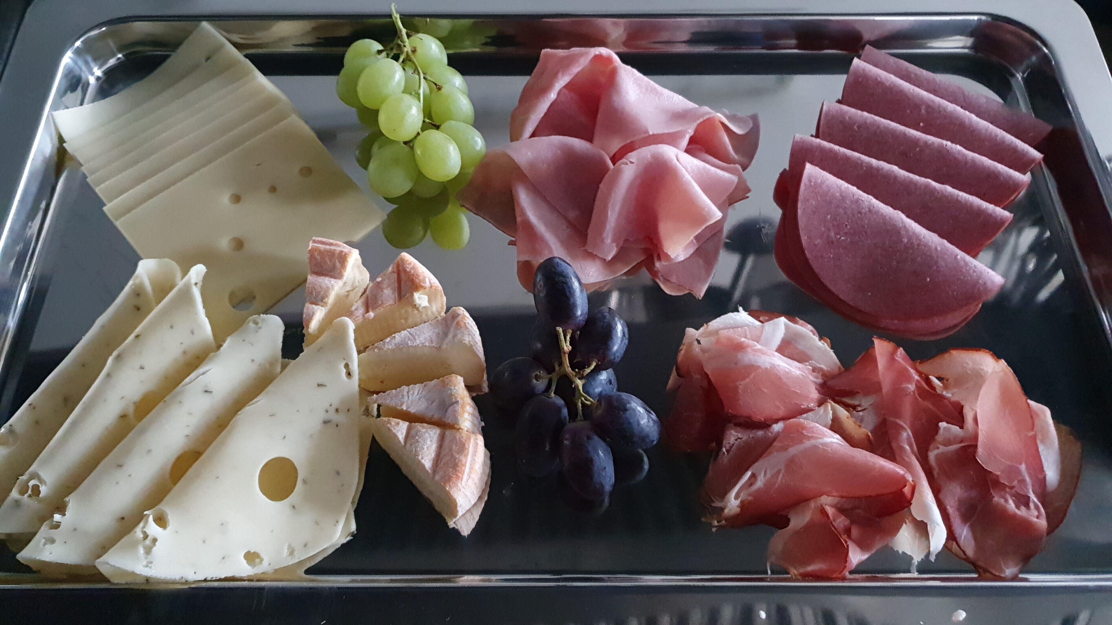 Uma bandeja com diversos alimentos, incluindo queijo, uvas e presunto.
