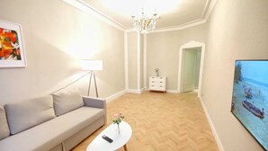 Uma sala de estar com um sofá, uma mesinha com um vaso, um abajur, uma televisão e um chão com padrão herringbone.