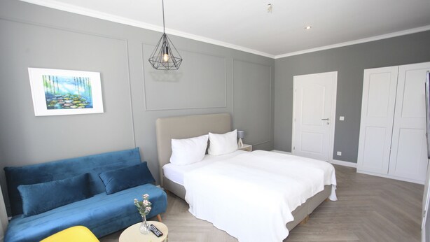 Quarto com cama, sofá azul, poltrona amarela, mesa redonda pequena com um vaso e um quadro na parede.