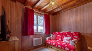 Um quarto acolhedor com paredes de madeira, um sofá vermelho com estampas de corações, um móvel de madeira para televisão e uma janela com cortinas vermelhas.