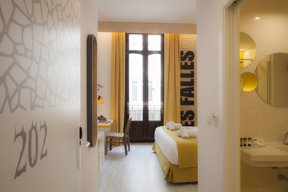 Quarto de hotel com uma cama, uma escrivaninha e uma cadeira. Há uma porta com um número.