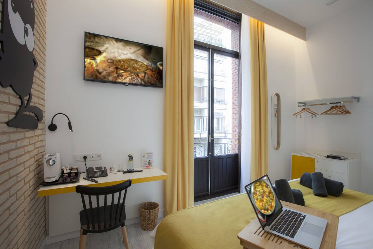 Quarto de hotel com escrivaninha, cadeira, cama e janela com cortinas.