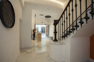 Um hall de hotel com um relógio de parede grande, uma mesa de recepção e uma escadaria com corrimãos ornamentados.
