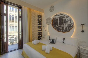 Quarto de hotel com uma cama, um espalha amarelo, almofadas brancas, um laptop na cama e uma parede grande com design circular e texto.