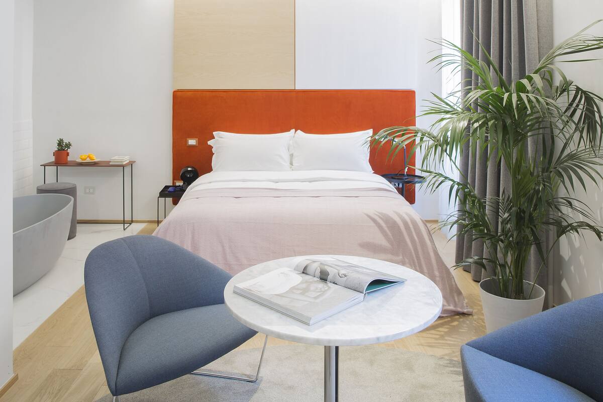 Quarto de hotel moderno com uma cama grande, duas poltronas azuis, uma mesinha redonda com uma revista, um vaso de planta e uma mesa de cabeceira com abajur.