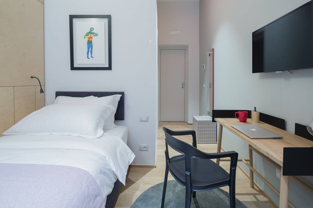 Quarto de hotel moderno com uma cama, uma escrivaninha com um laptop, uma televisão e um quadro na parede.