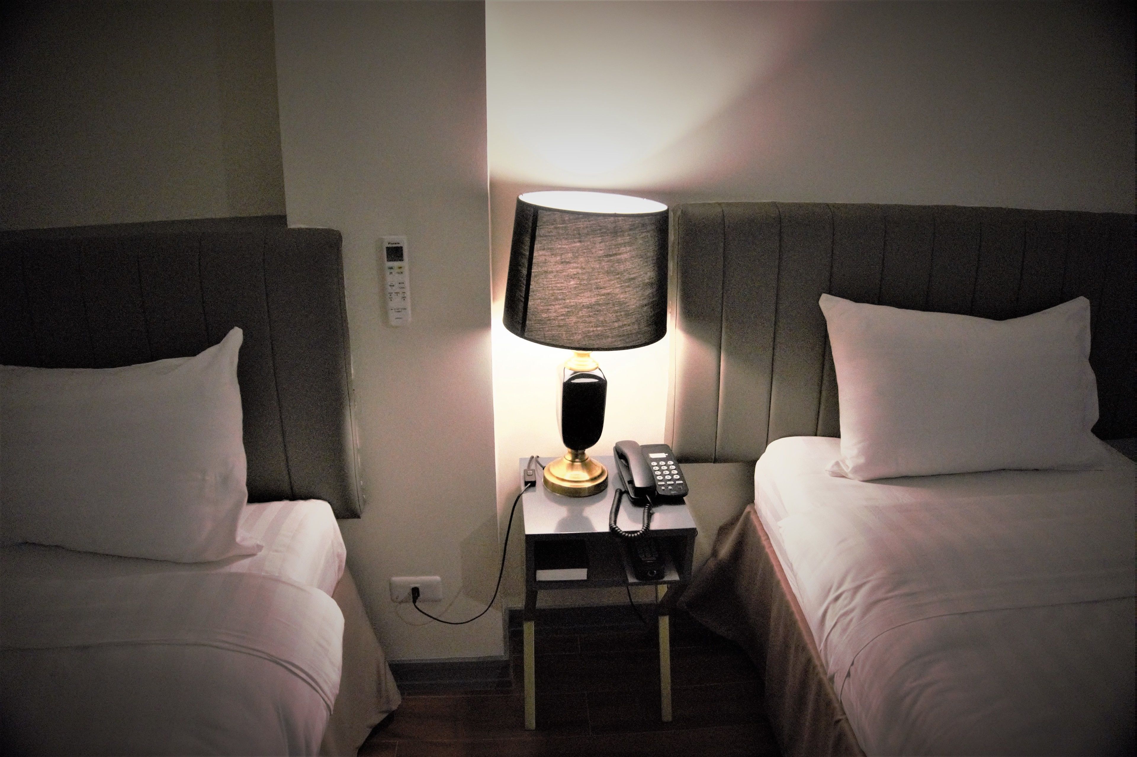 Quarto de hotel com duas camas de solteiro, mesa de cabeceira com luminária, telefone e tomada.