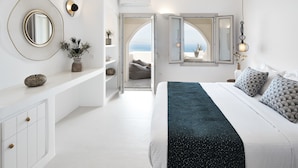 Quarto moderno com uma cama grande, mesa de cabeceira, espelho e vista para o mar através de uma porta aberta.