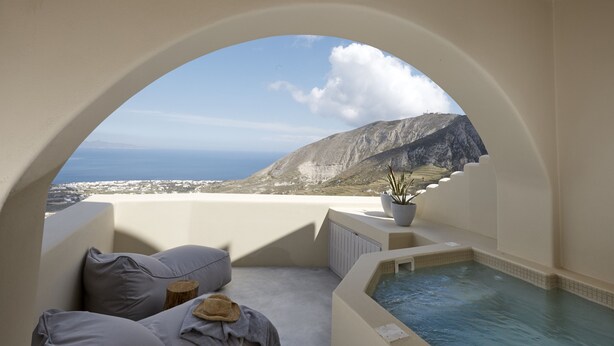 Uma varanda com vista para o mar e montanhas, uma banheira de hidromassagem e um espaço de estar com almofadas e um chapéu.