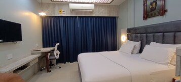 Quarto de hotel com cama, escrivaninha, cadeira e televisão na parede.
