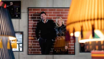 Um homem e uma mulher em frente a um quadro com parede de tijolos ao fundo.