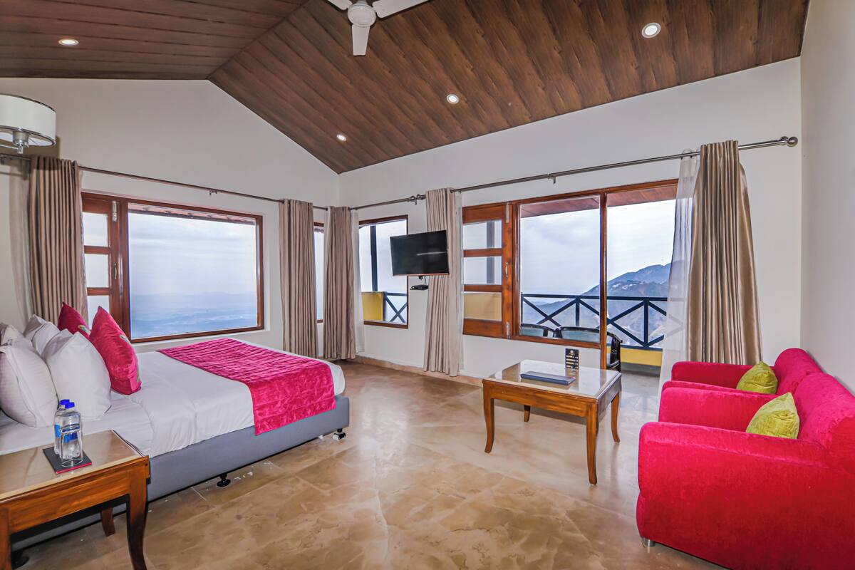 Quarto de hotel com cama, sofá vermelho, televisão e amplas janelas com vista.