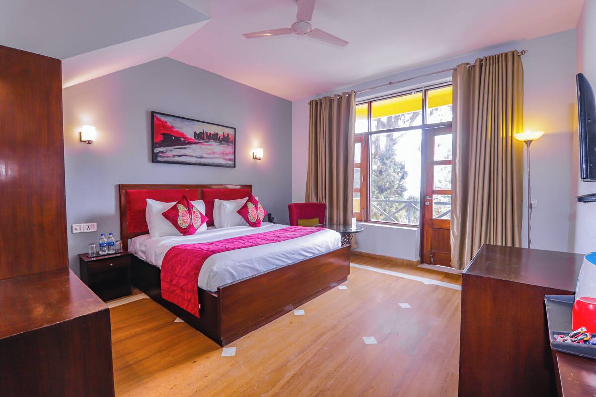 Quarto de hotel com uma cama grande, cabeceira de madeira, mesa de cabeceira, escrivaninha, cadeira, janela com cortinas, quadro na parede e ventilador de teto.