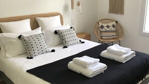 Cama bem arrumada com almofadas brancas e estampadas, manta preta e toalhas dobradas. Há uma cadeira de vime com almofada estampada, um painel de macramê fixado na parede e um lustre pendente.