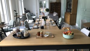 Uma sala de jantar com uma longa mesa de madeira posta para uma refeição, rodeada de cadeiras, com vista para o exterior através de grandes janelas.