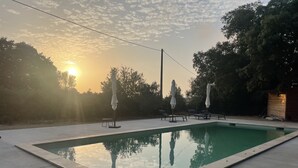 Piscina retangular com espreguiçadeiras e guarda-sóis, cercada por um terraço ladrilhado, com árvores e um pôr do sol ao fundo.